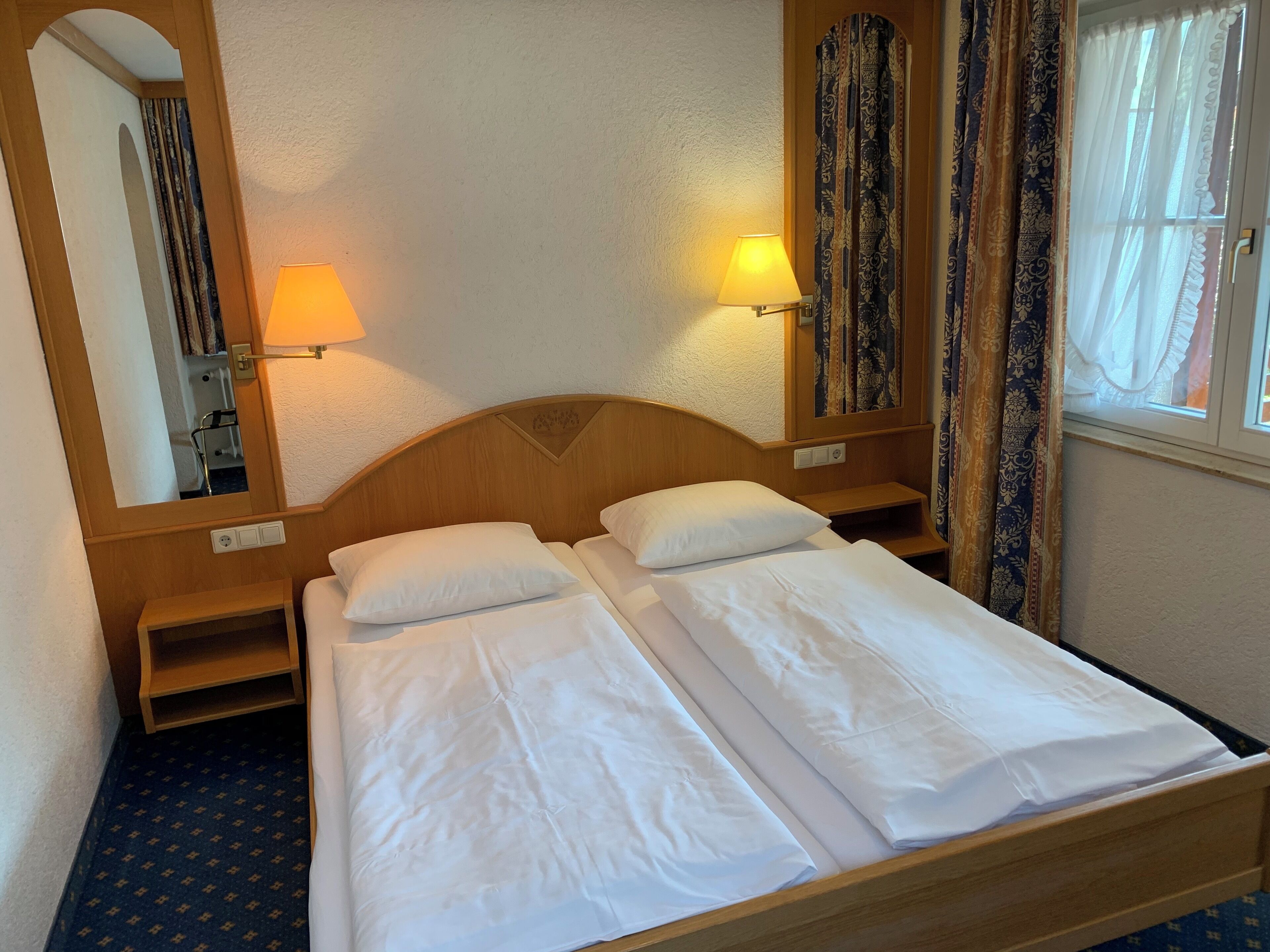 Uma cama de casal com roupa de cama branca, duas mesas de cabeceira, um espelho e duas luminárias fixadas na parede.