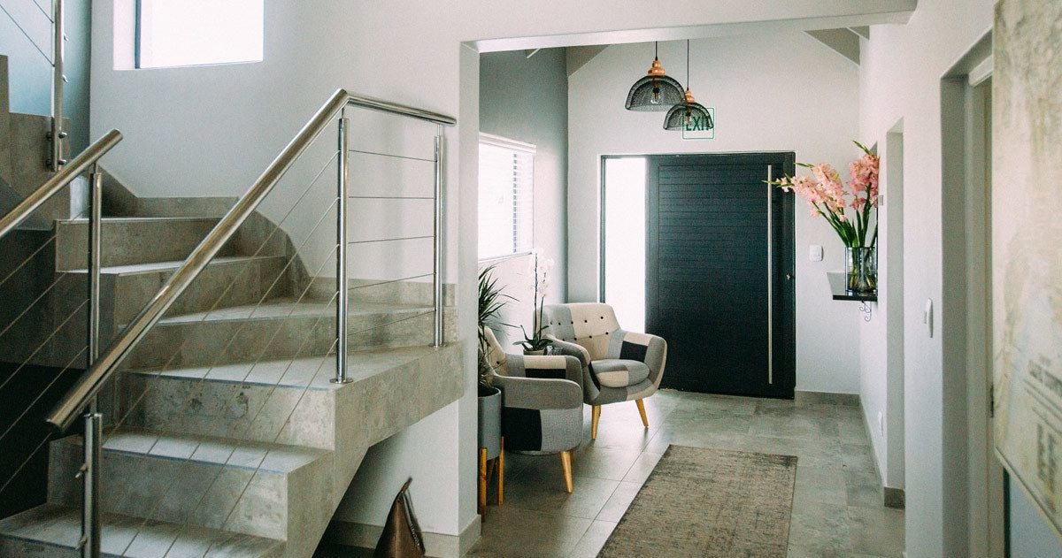 Um interior moderno com escada, área de estar com duas poltronas, porta escura e luminária suspensa.