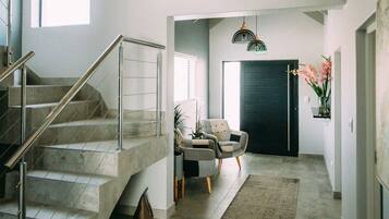 Um interior moderno com escada, área de estar com duas poltronas, porta escura e luminária suspensa.