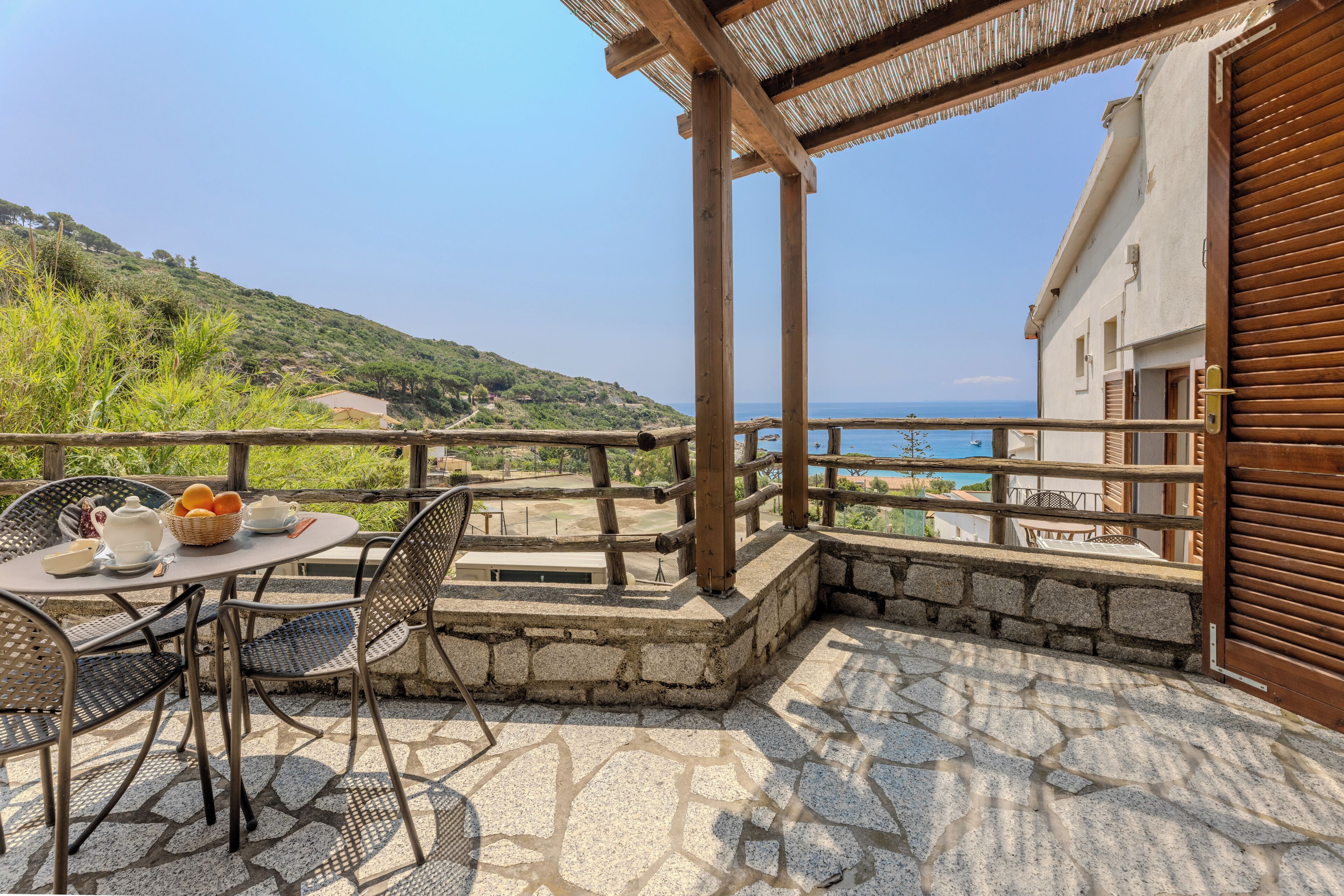 Um terraço com piso de pedra, cadeiras de metal, uma mesa com um bule e uma cesta de frutas, e vista para o mar e as colinas.