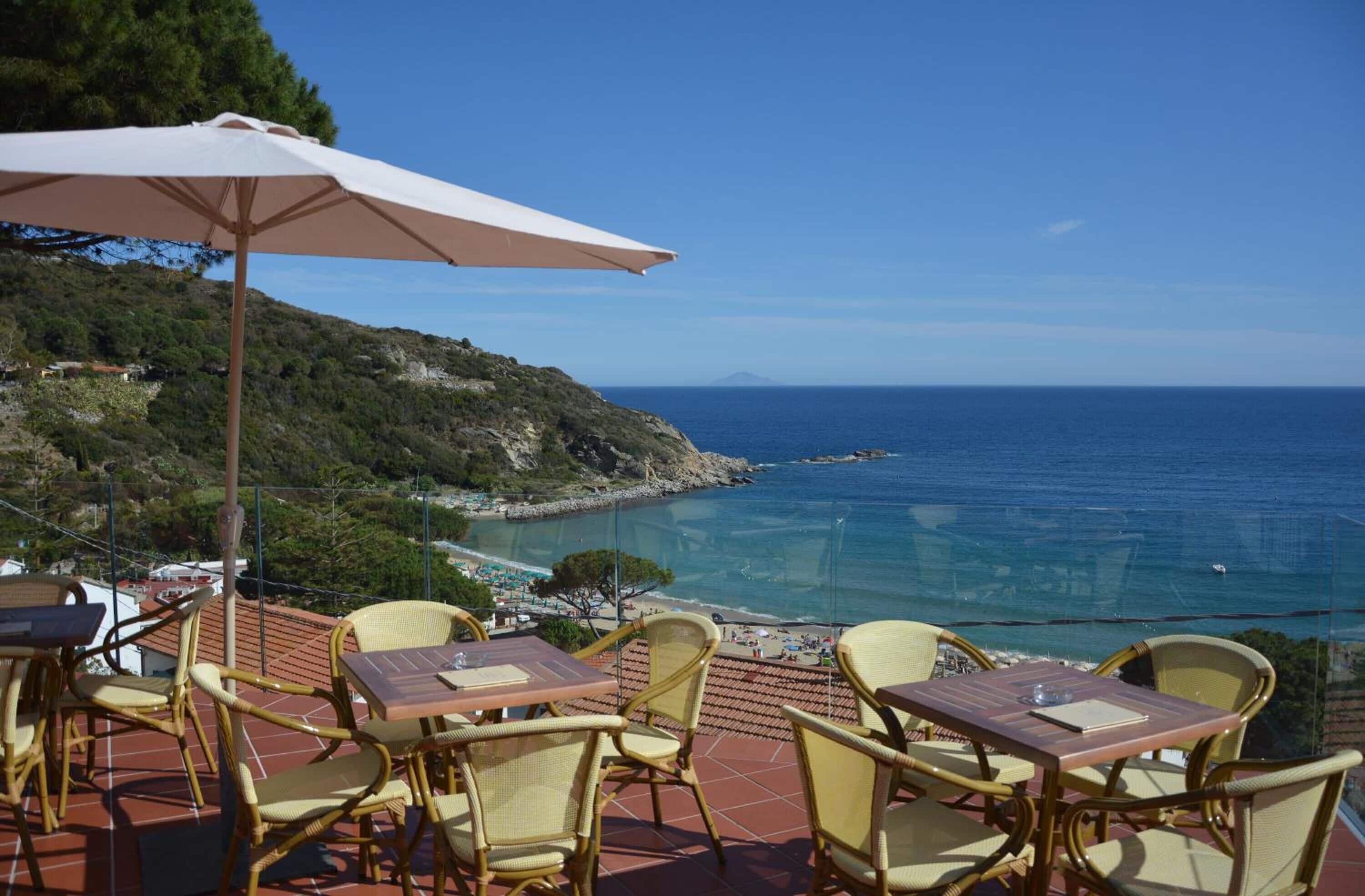 Terraço ao ar livre com cadeiras e mesas de vime, com vista para a paisagem costeira, mar azul cristalino e montanhas ao longe.