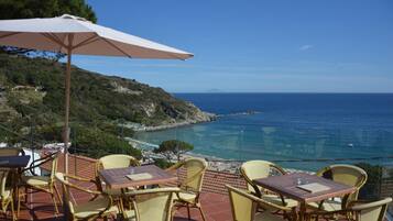 Terraço ao ar livre com cadeiras e mesas de vime, com vista para a paisagem costeira, mar azul cristalino e montanhas ao longe.