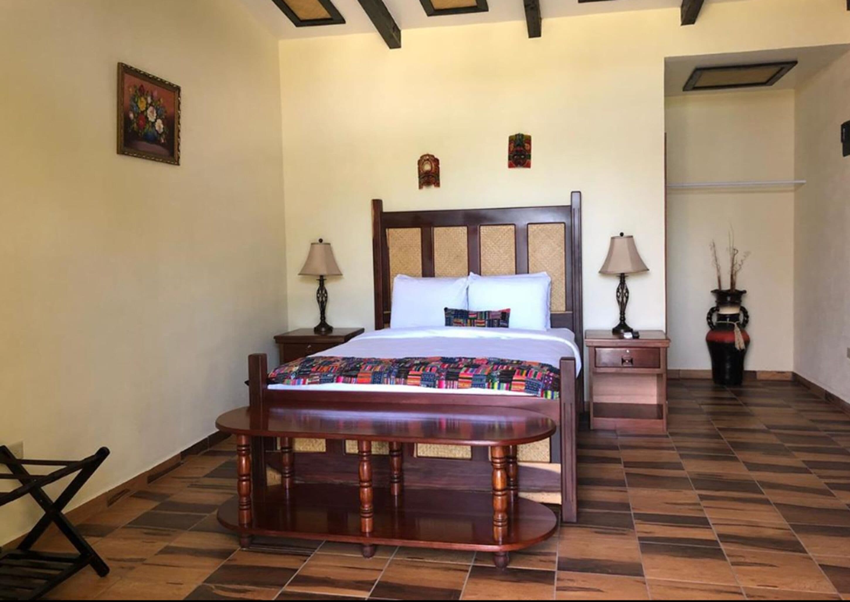 Quarto de hotel com uma cama grande, duas mesas de cabeceira com abajures, um banquinho e um vaso de planta.
