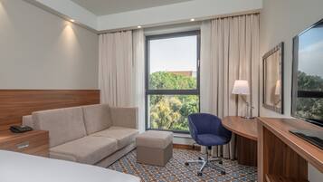Quarto de hotel com sofá, banquinho, escrivaninha com luminária, televisão e uma grande janela com cortinas.
