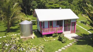 Uma casa pequena com telhado branco e detalhes em rosa, cercada por vegetação exuberante e um caminho de pedra.