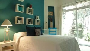 Quarto com cama, mesas de cabeceira brancas, paredes verde-água e janela com vista para árvores.