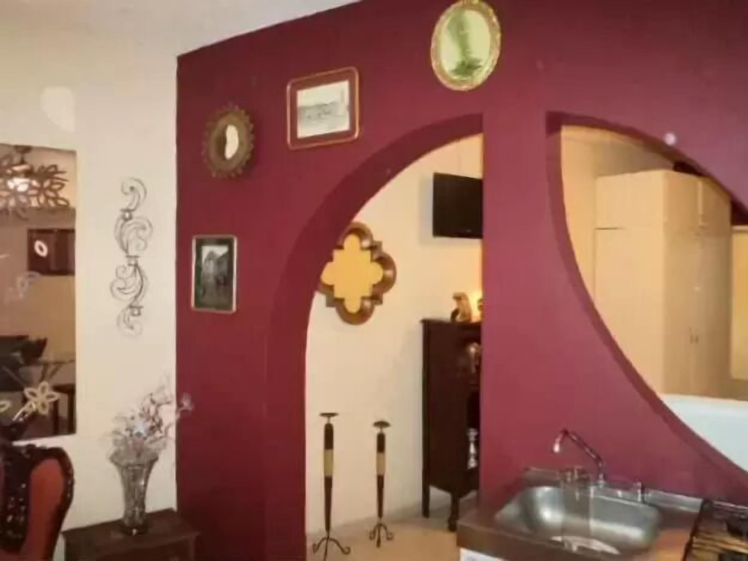 Quarto com parede em vermelho, um espelho e um quadro emoldurado. Há um vaso com flores sobre uma mesa e um suporte de metal.