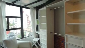 Quarto com sofá branco, uma mesinha pequena e uma janela grande. Há também um guarda-roupa branco com várias gavetas e uma porta.