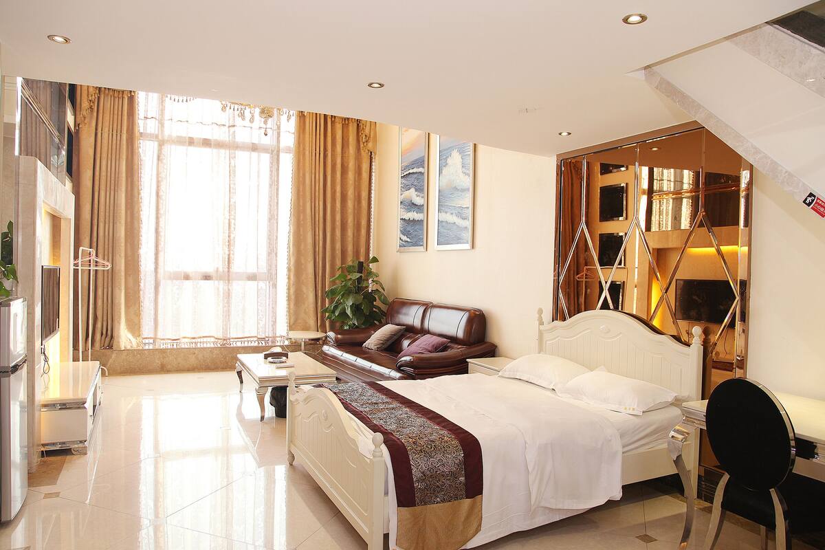 Quarto de hotel com cama, escrivaninha, cadeira, televisão, planta e uma janela ampla com cortinas.