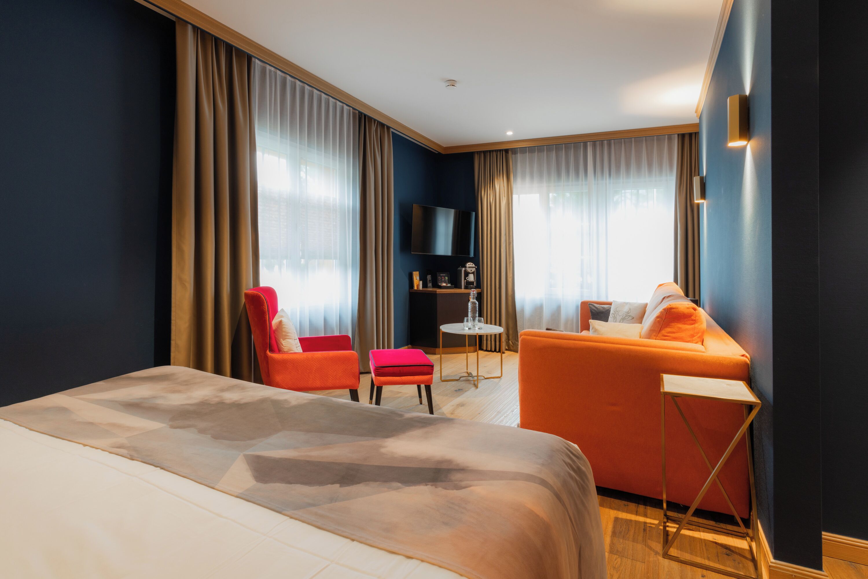 Quarto de hotel com cama, sofá laranja, poltrona vermelha e uma pequena mesa.