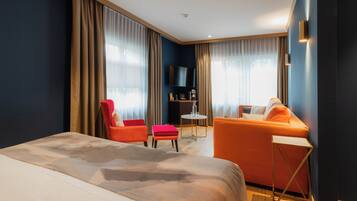 Quarto de hotel com cama, sofá laranja, poltrona vermelha e uma pequena mesa.