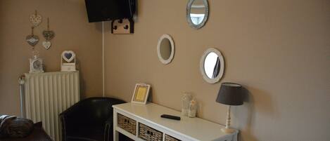 Quarto com cama, escrivaninha com cestos, cadeira, televisão e espelhos na parede.