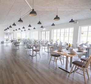 Uma sala de jantar espaçosa com várias mesas e cadeiras, janelas amplas e iluminação pendente.