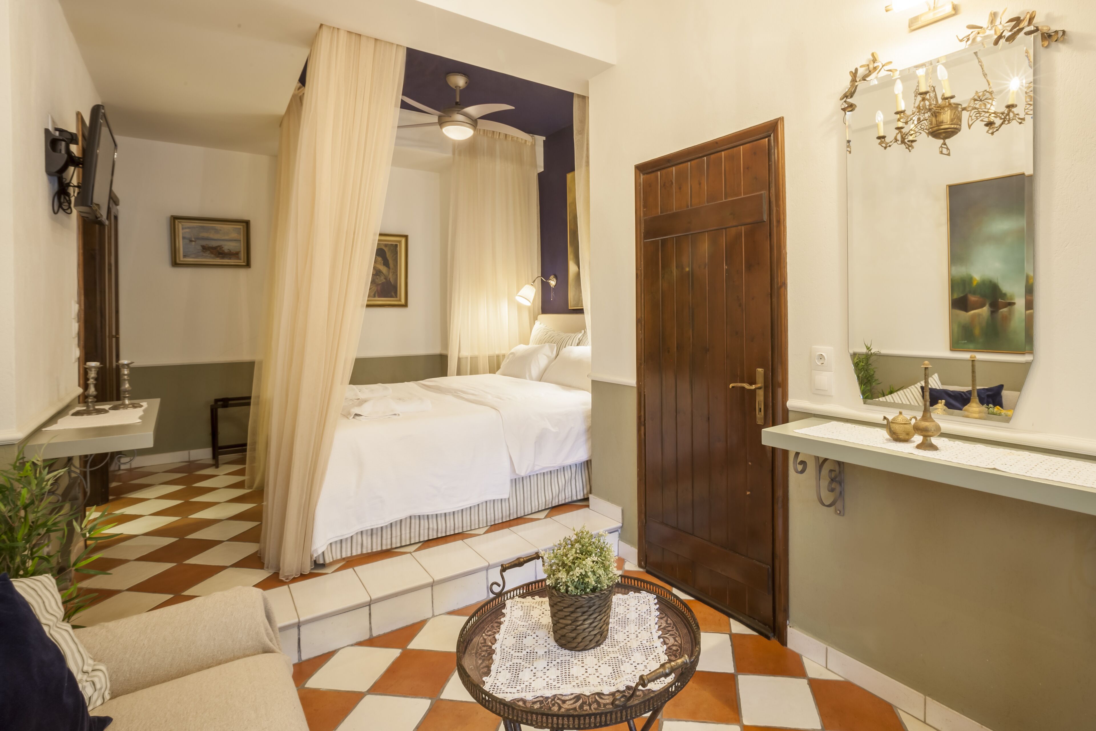 Quarto com cama, mesa de cabeceira, cadeira, pequena mesa com planta, ventilador de teto, espelho e porta.