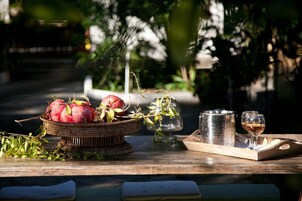 Uma mesa de madeira com uma cesta de vime contendo frutas, um jarro de metal e um copo de vinho.