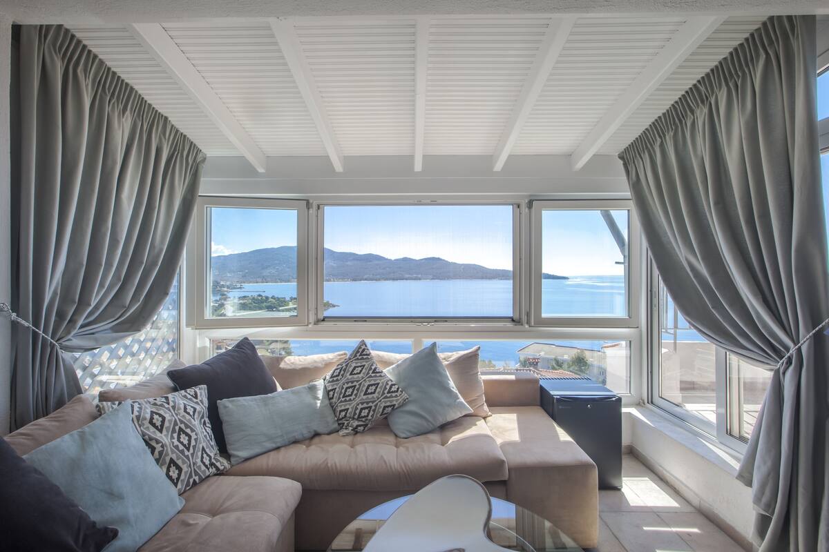 Uma sala de estar moderna com um sofá bege, almofadas estampadas e vista para o mar através de amplas janelas.