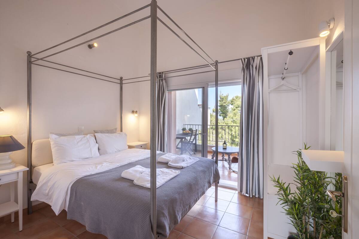 Uma cama de dossel com lençóis brancos, uma mesa de cabeceira com abajur e uma varanda com mesa e cadeiras.