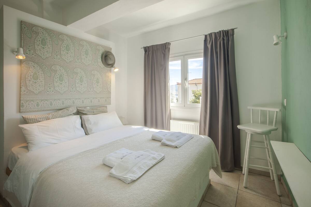 Cama bem arrumada com roupas de cama e toalhas brancas, uma cadeira branca e uma janela com cortinas.
