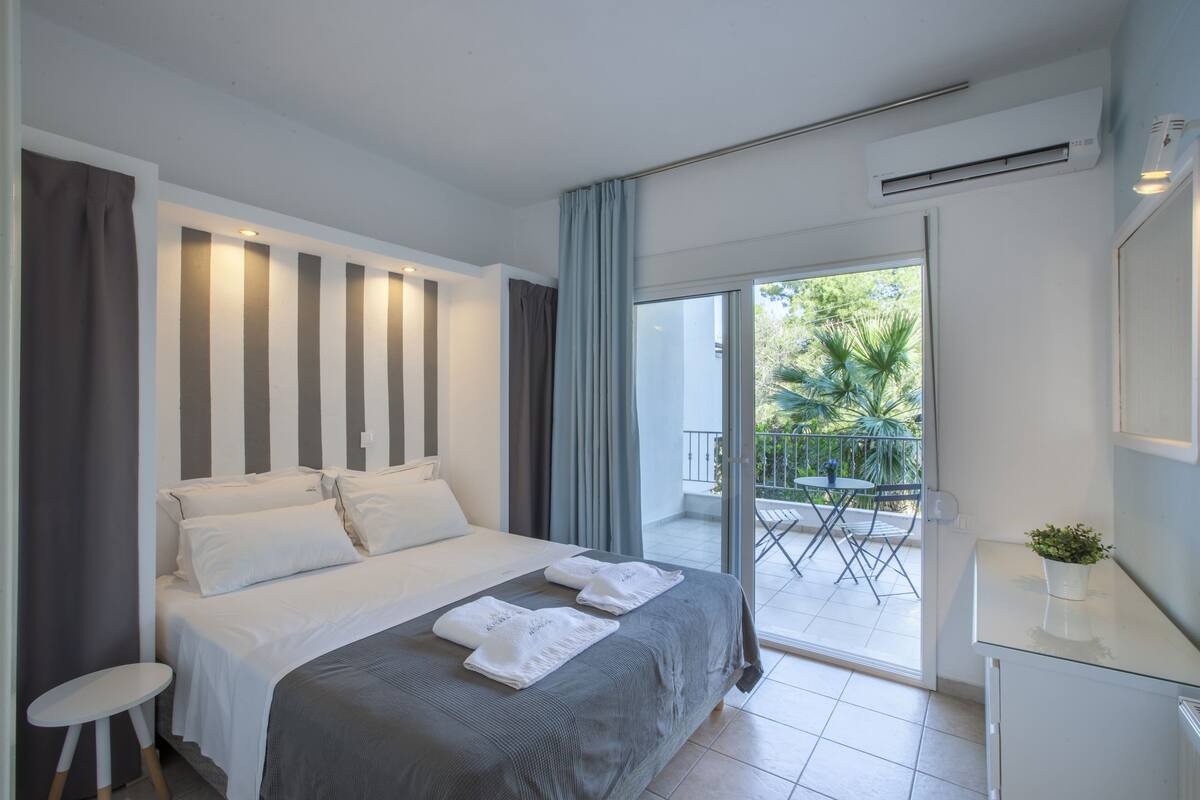 Quarto de hotel moderno com uma cama grande, uma mesa redonda pequena, uma planta em vaso e uma varanda com vista para áreas verdes.