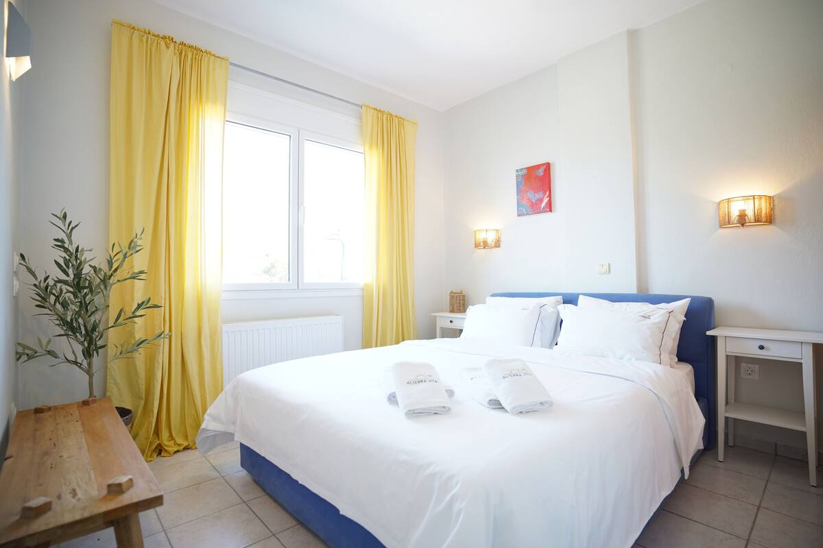 Quarto de hotel com cama, mesa de cabeceira, cortinas amarelas na janela, luminária na parede e planta em vaso.
