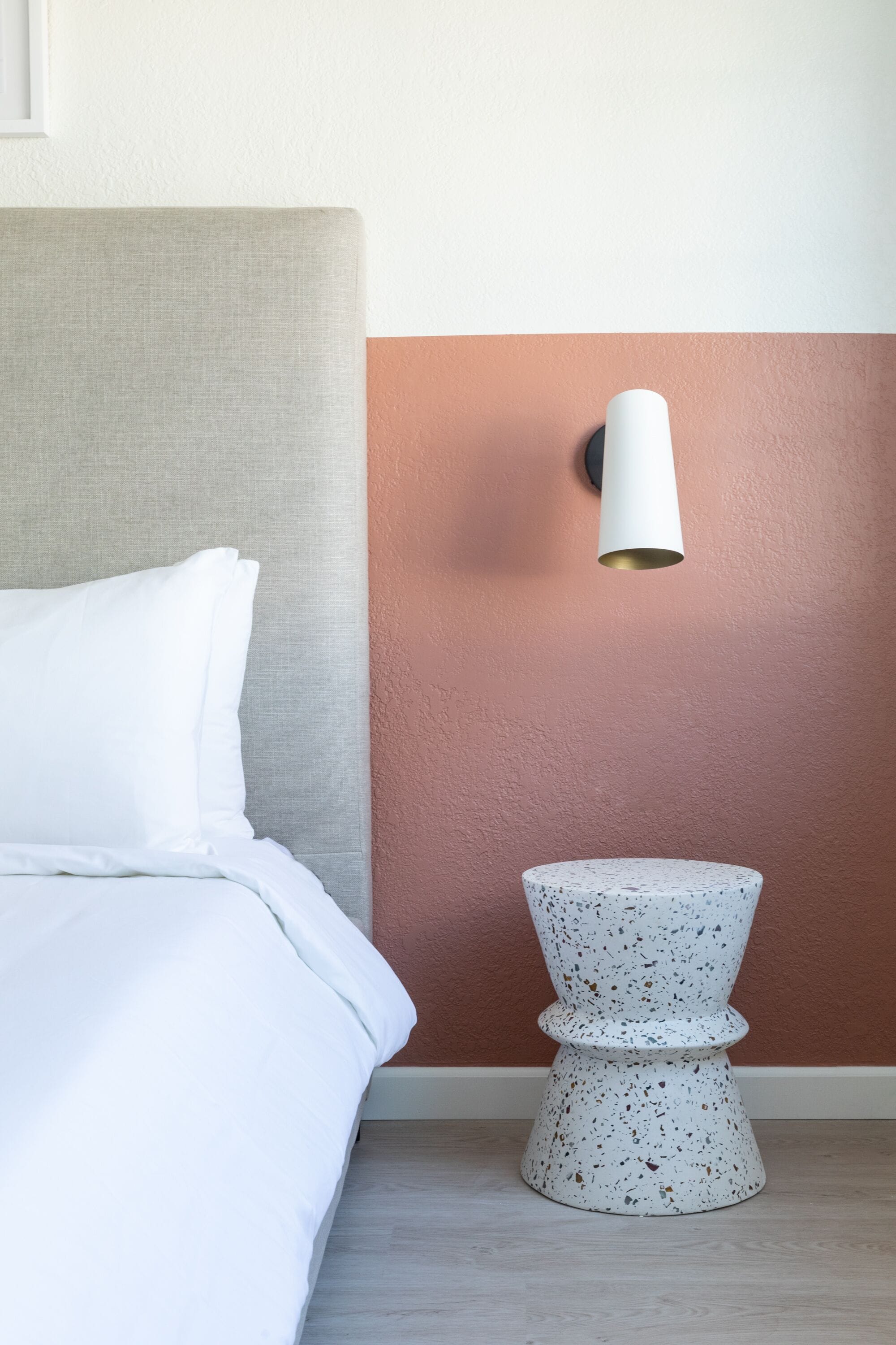 Quarto de hotel com cama, luminária fixada na parede e mesa de cabeceira com padrão salpicado.