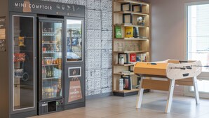 Uma mini-cozinha com geladeira abastecida com bebidas e lanches, uma estante de madeira com livros e objetos decorativos, e uma mesa de pebolim.