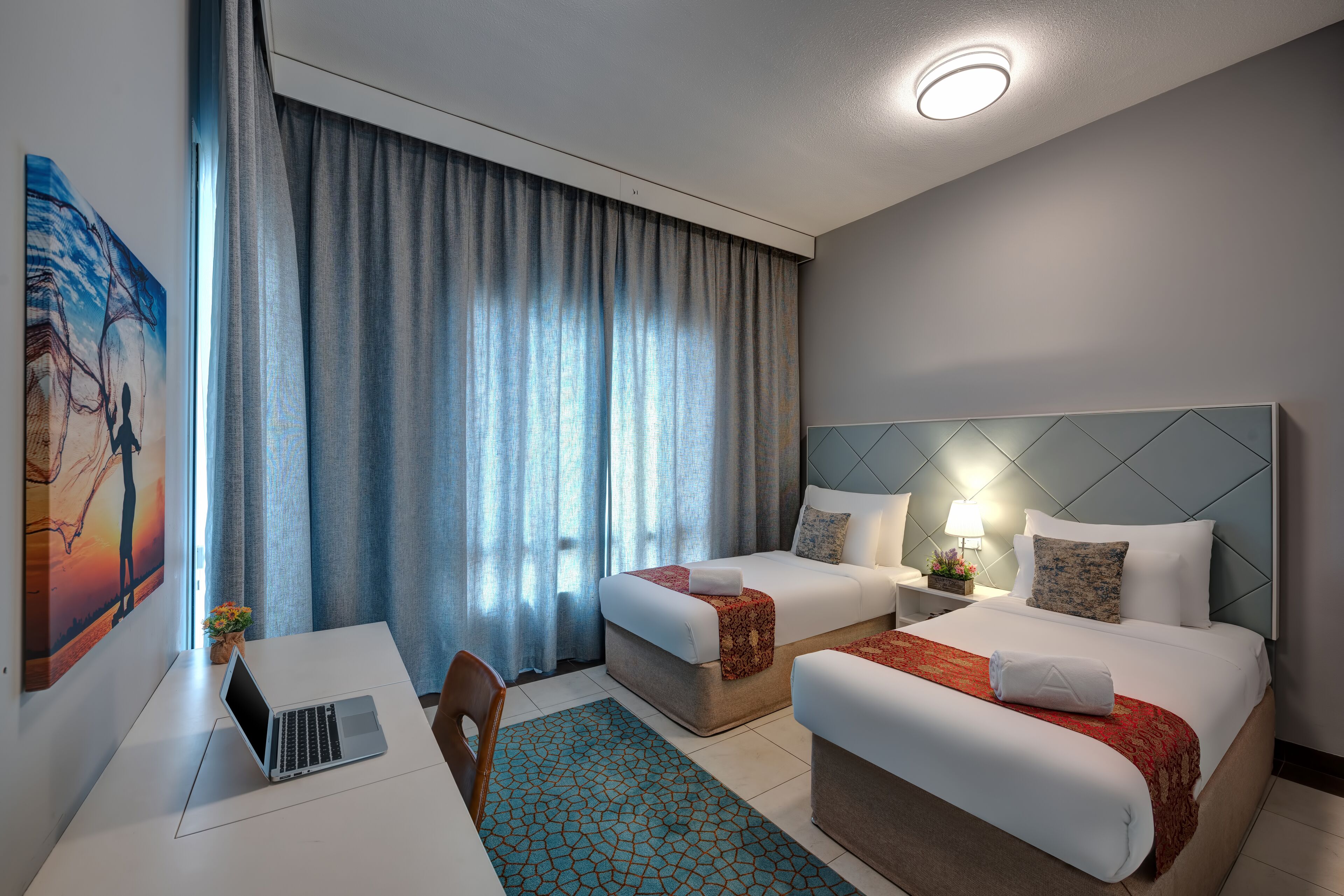 Quarto de hotel com duas camas, uma secretária com um laptop, um quadro na parede e uma janela grande com cortinas.