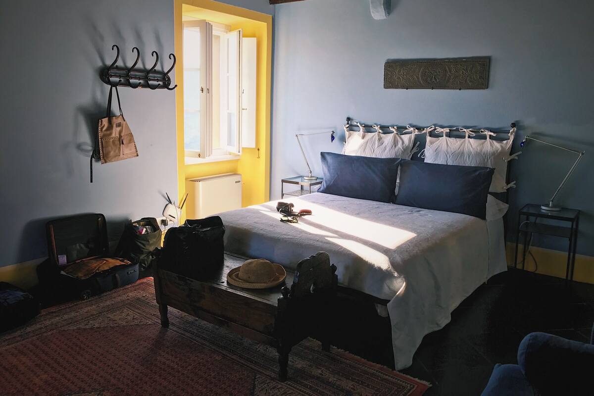 Quarto com cama, mesa de cabeceira, abajur e janela com cortinas.