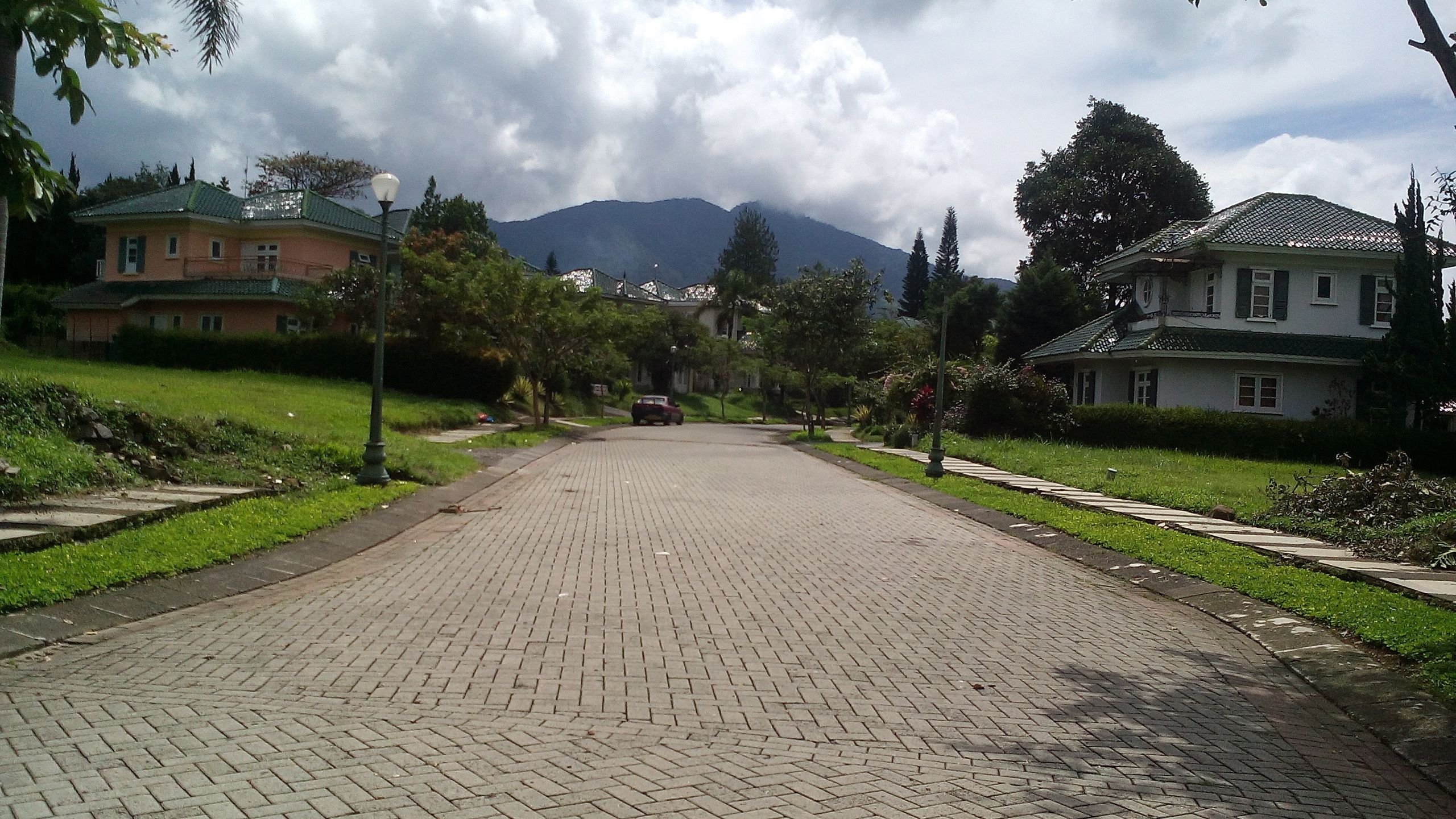 Rua pavimentada com casas, um carro e uma montanha ao fundo.