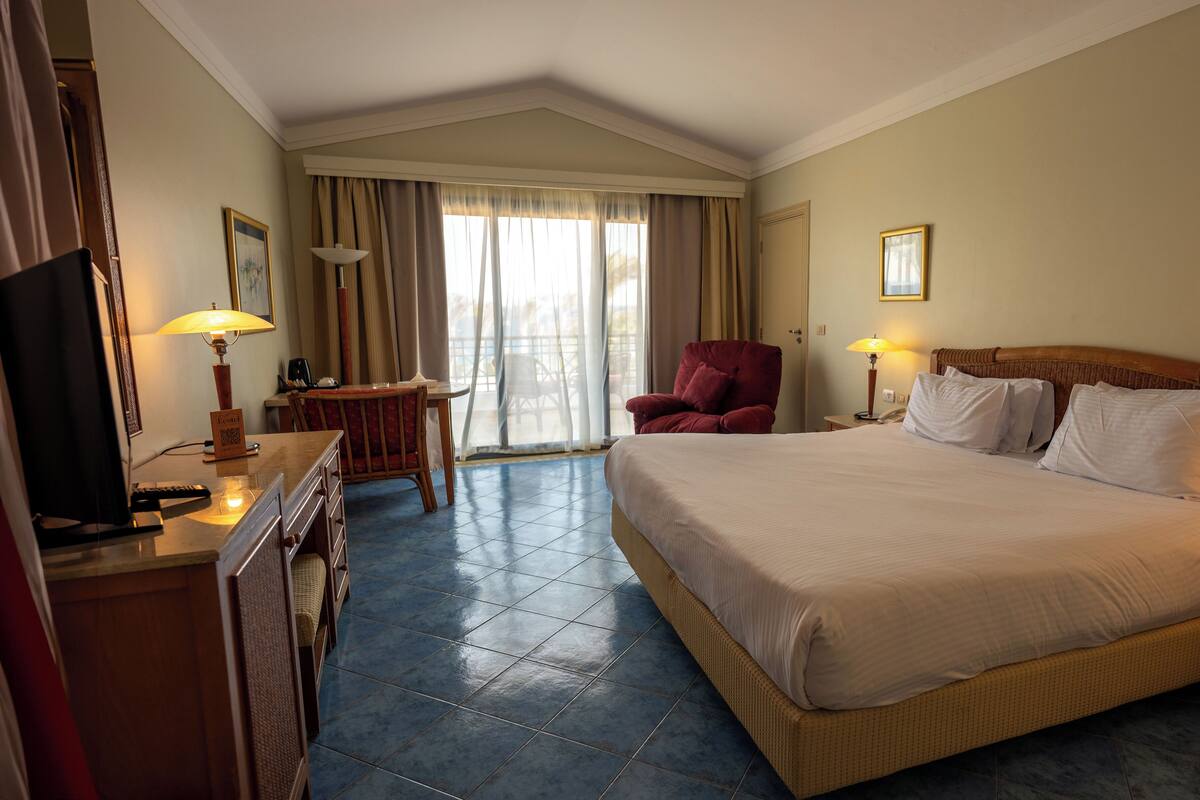 Quarto de hotel com uma cama grande, uma escrivaninha com candeeiro, uma cadeira e uma televisão.