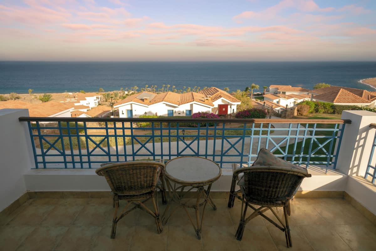 Varanda com cadeiras de vime e uma pequena mesa, com vista para uma vila costeira com casas e o mar.