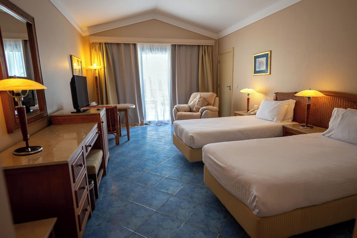 Quarto de hotel com duas camas, uma escrivaninha com cadeira, televisão e uma janela grande com cortinas.