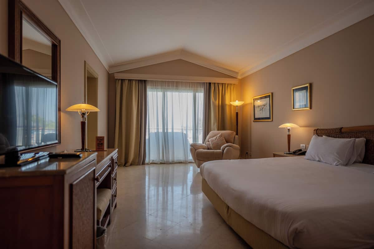 Quarto de hotel com uma cama grande, uma escrivaninha com luminária, uma cadeira e vista para o exterior pela janela.