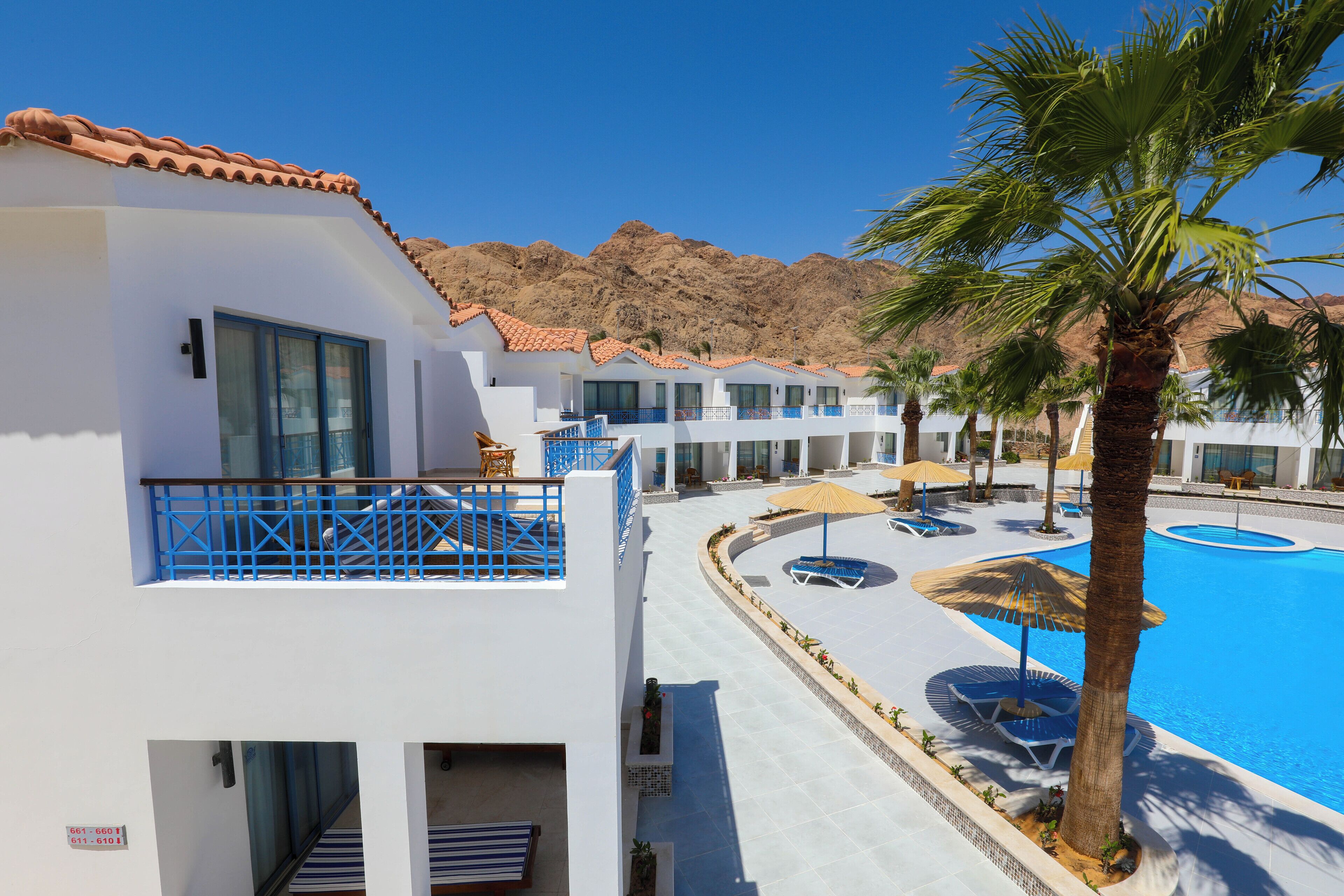 Resort com piscina, palmeiras e montanhas ao fundo.