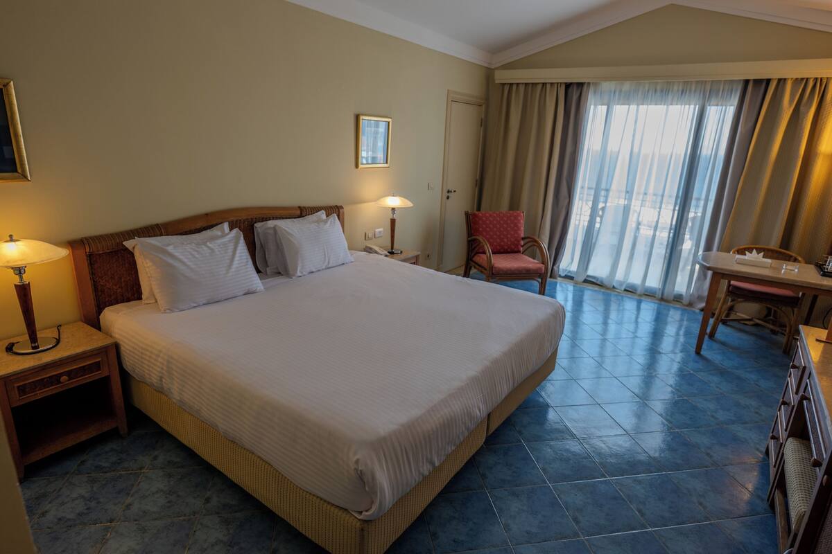 Quarto de hotel com uma cama grande, uma cadeira, uma escrivaninha e uma janela com cortinas.