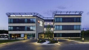 Um hotel moderno com restaurante, fachada de vidro, área externa para sentar e entrada ajardinada.