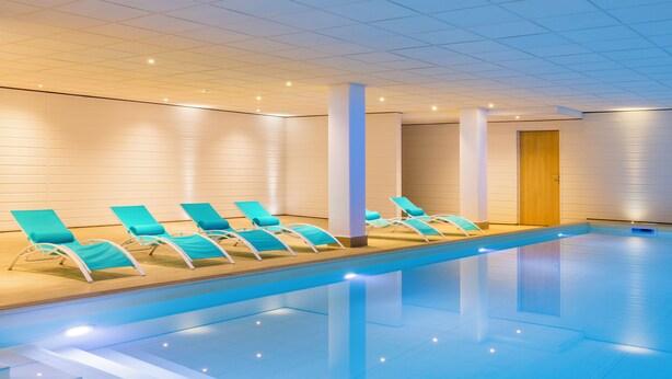 Uma piscina interior com água azul, rodeada por paredes e teto brancos, com várias espreguiçadeiras turquesa dispostas ao longo da borda da piscina.