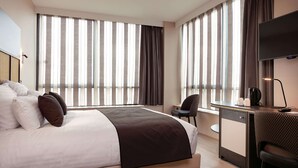 Quarto de hotel com uma cama grande, uma escrivaninha, uma cadeira, televisão de tela plana e janelas amplas com cortinas.