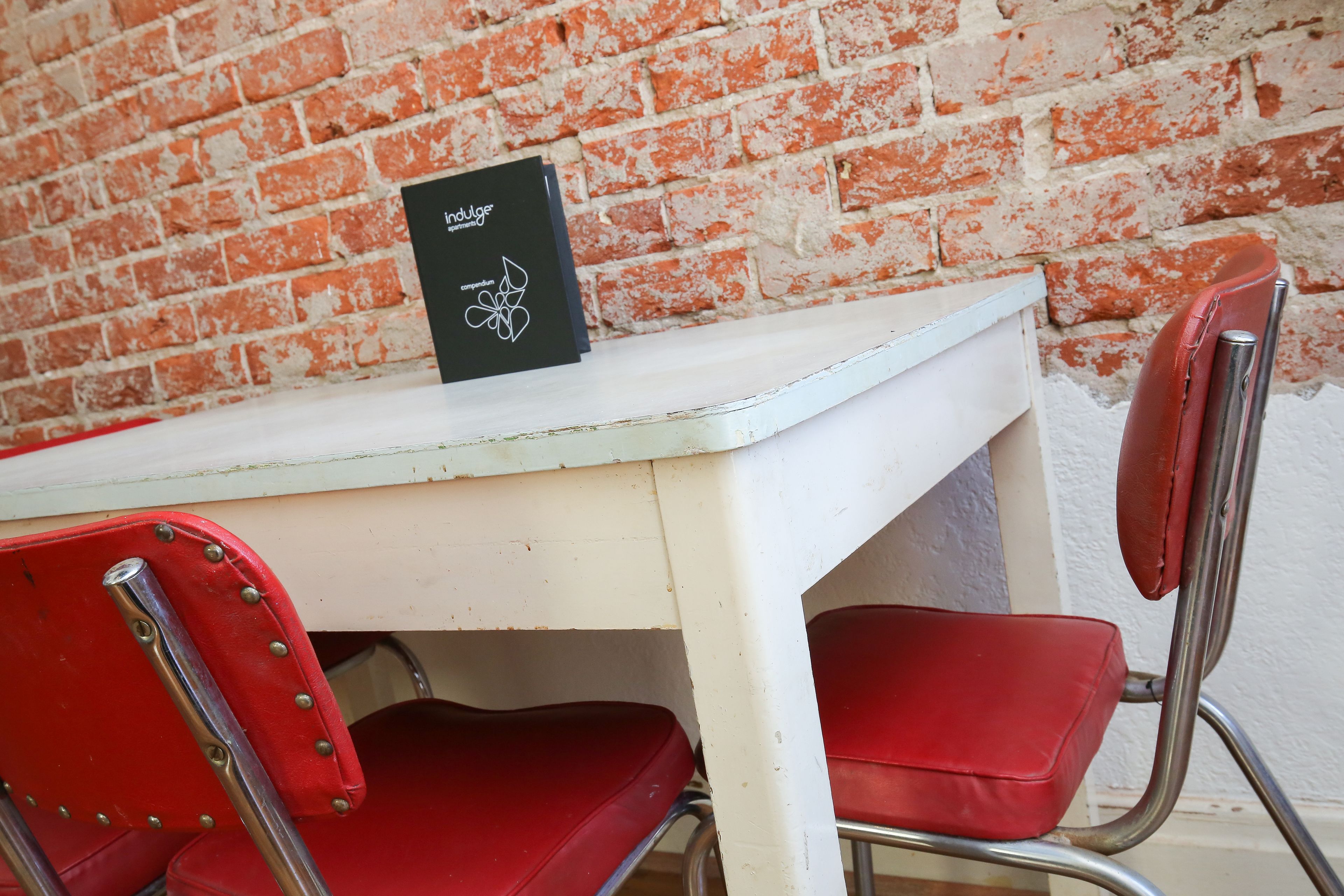 Uma mesa branca com um livro preto intitulado 'indulge' em frente a uma parede de tijolos.