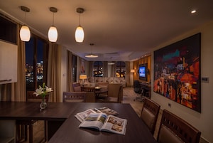 Quarto de hotel com área de refeições, um conjunto de sofás, televisão e uma grande pintura na parede.