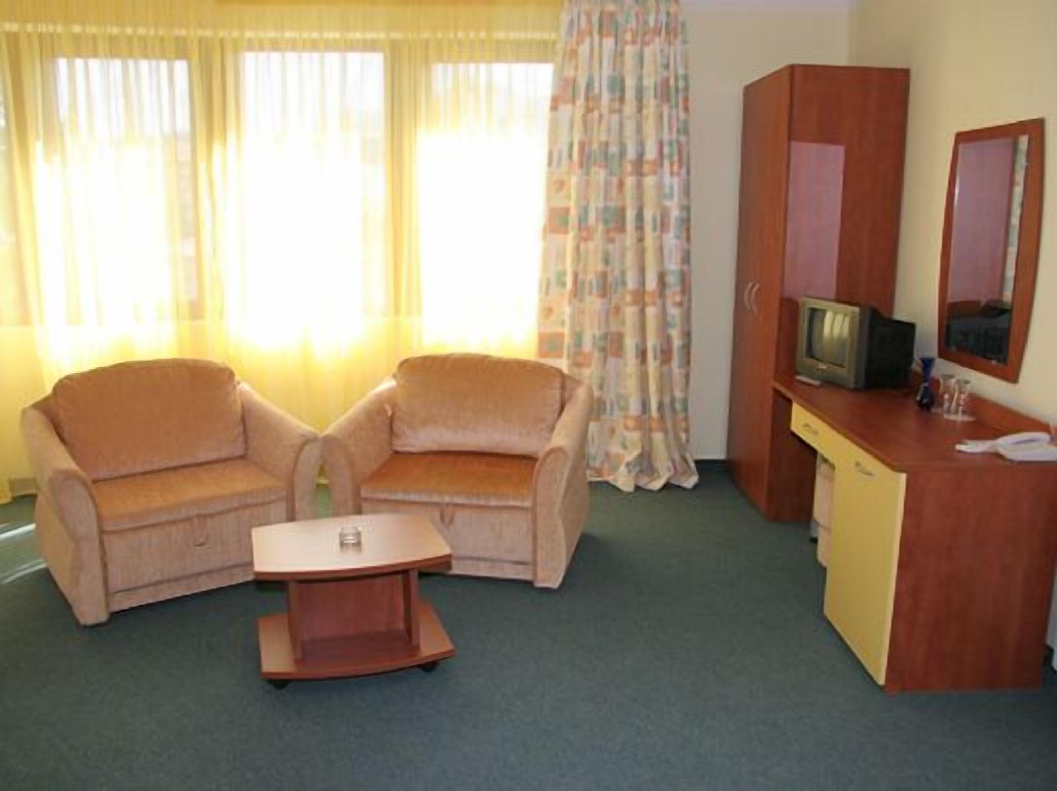 Quarto de hotel com duas poltronas, uma pequena mesa de madeira, uma televisão em um móvel de madeira e um espelho.