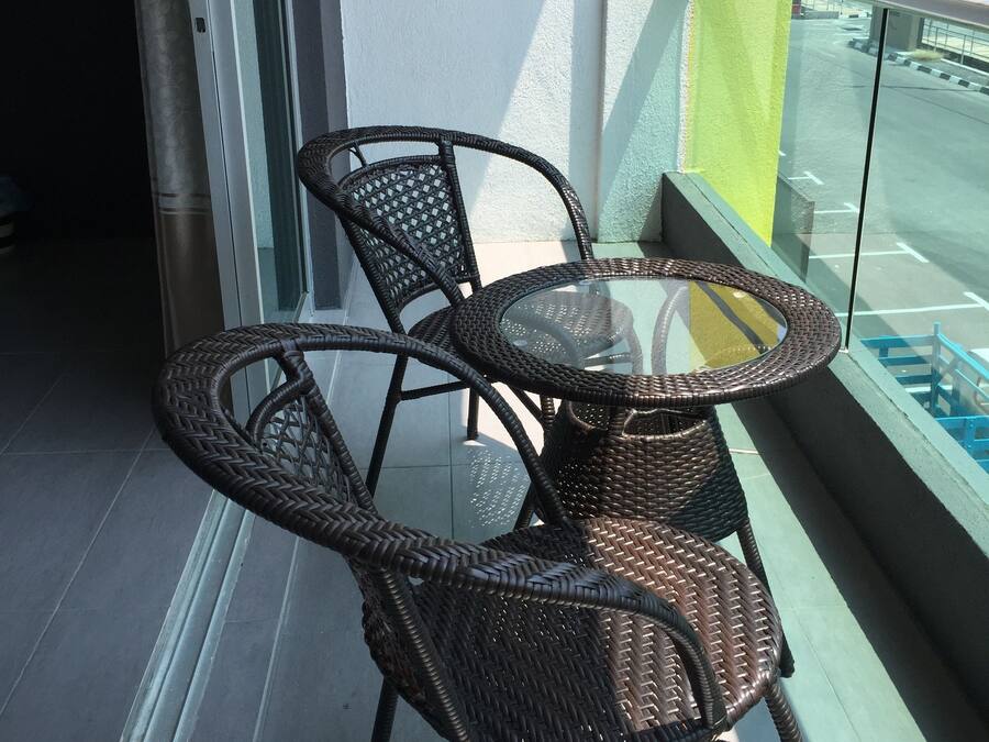 Varanda com cadeiras de vime e uma mesa de vidro, com vista para uma rua com edifícios e vegetação.