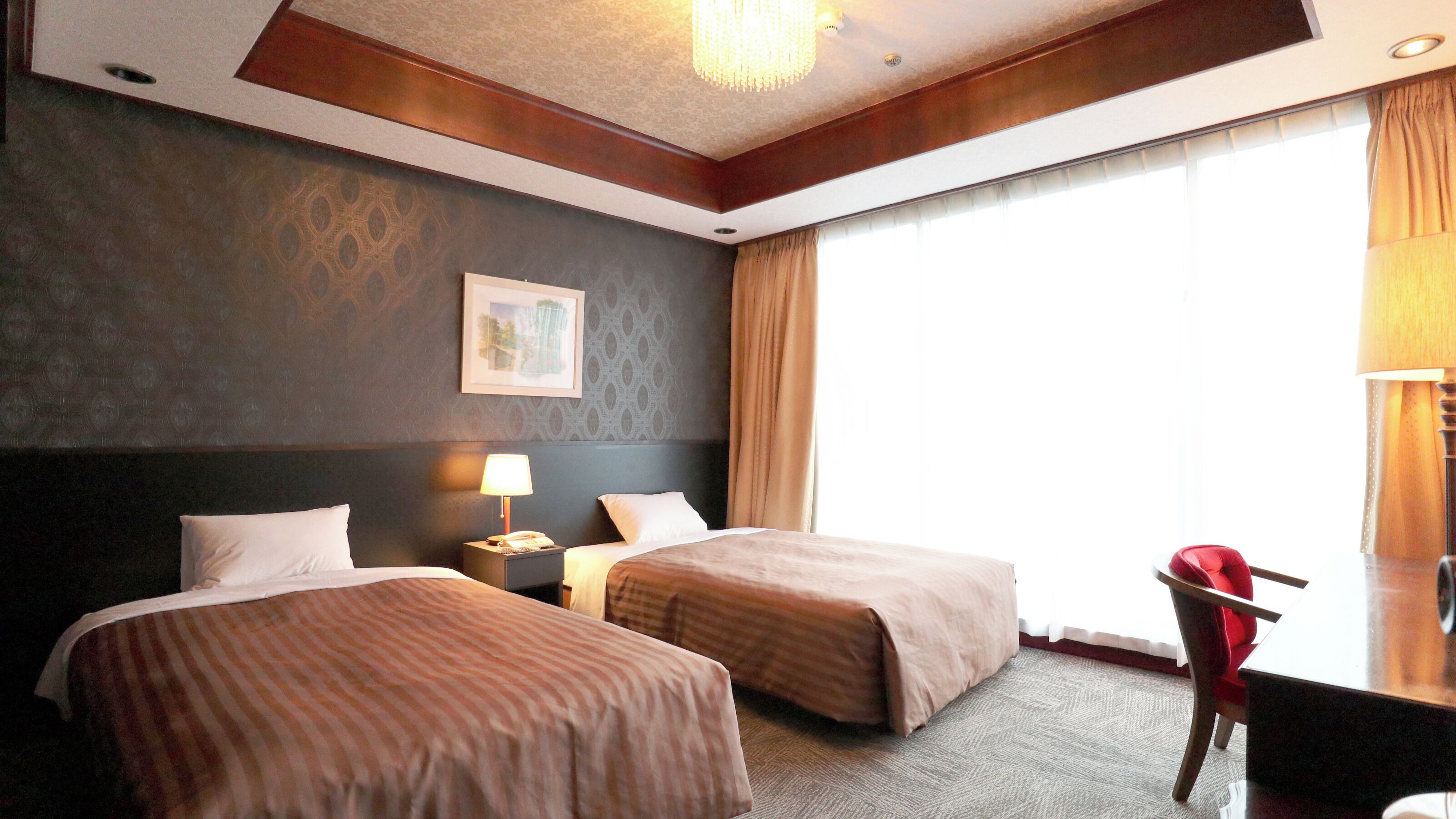 Quarto de hotel com duas camas, uma escrivaninha, uma cadeira, um abajur e uma janela grande.