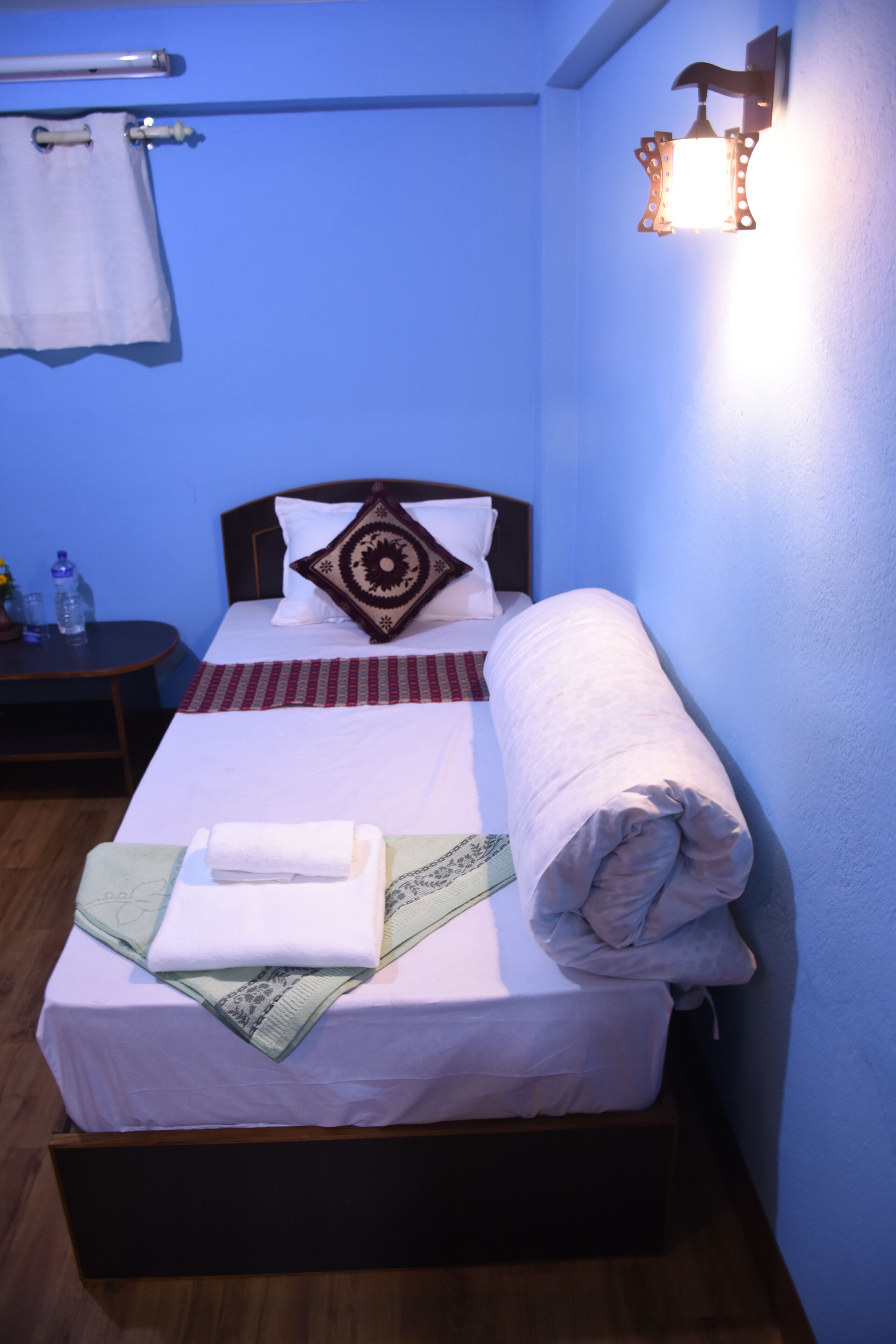 Cama bem arrumada com colcha branca, toalha dobrada e almofada decorativa. Há uma pequena mesa com uma garrafa e copos ao lado da cama.