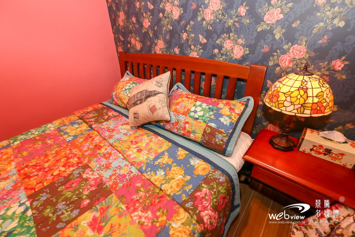 Quarto com colcha estampada de flores, cabeceira de madeira e mesa de cabeceira com abajur e uma caixa.