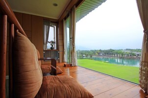 Quarto com grande janela que oferece vista para um lago e vegetação. Piso de madeira, sofá marrom e uma mesa pequena com um vaso.