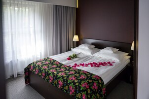 Uma cama bem arrumada com lençóis brancos, uma colcha floral e pétalas de rosa sobre a cama. Há uma mesa de cabeceira com uma luminária e um relógio.