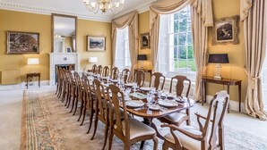Uma sala de jantar formal com uma longa mesa posta para refeição, cercada por cadeiras.