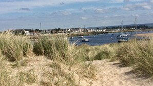 Cena costeira com dunas de areia, um porto com barcos e edifícios ao fundo.
