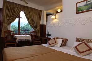 Quarto de hotel com uma cama grande, duas cadeiras, uma mesa e vista para o verde pela janela.